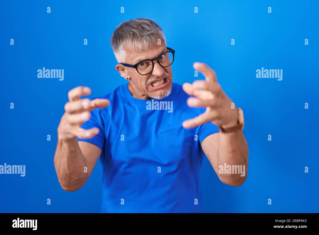 Hispanic man with grey hair standing over blue background shouting ...