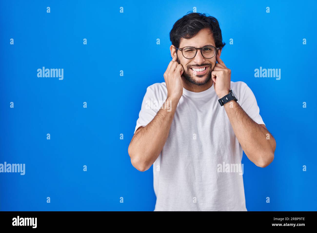 Handsome latin man standing over blue background covering ears with ...