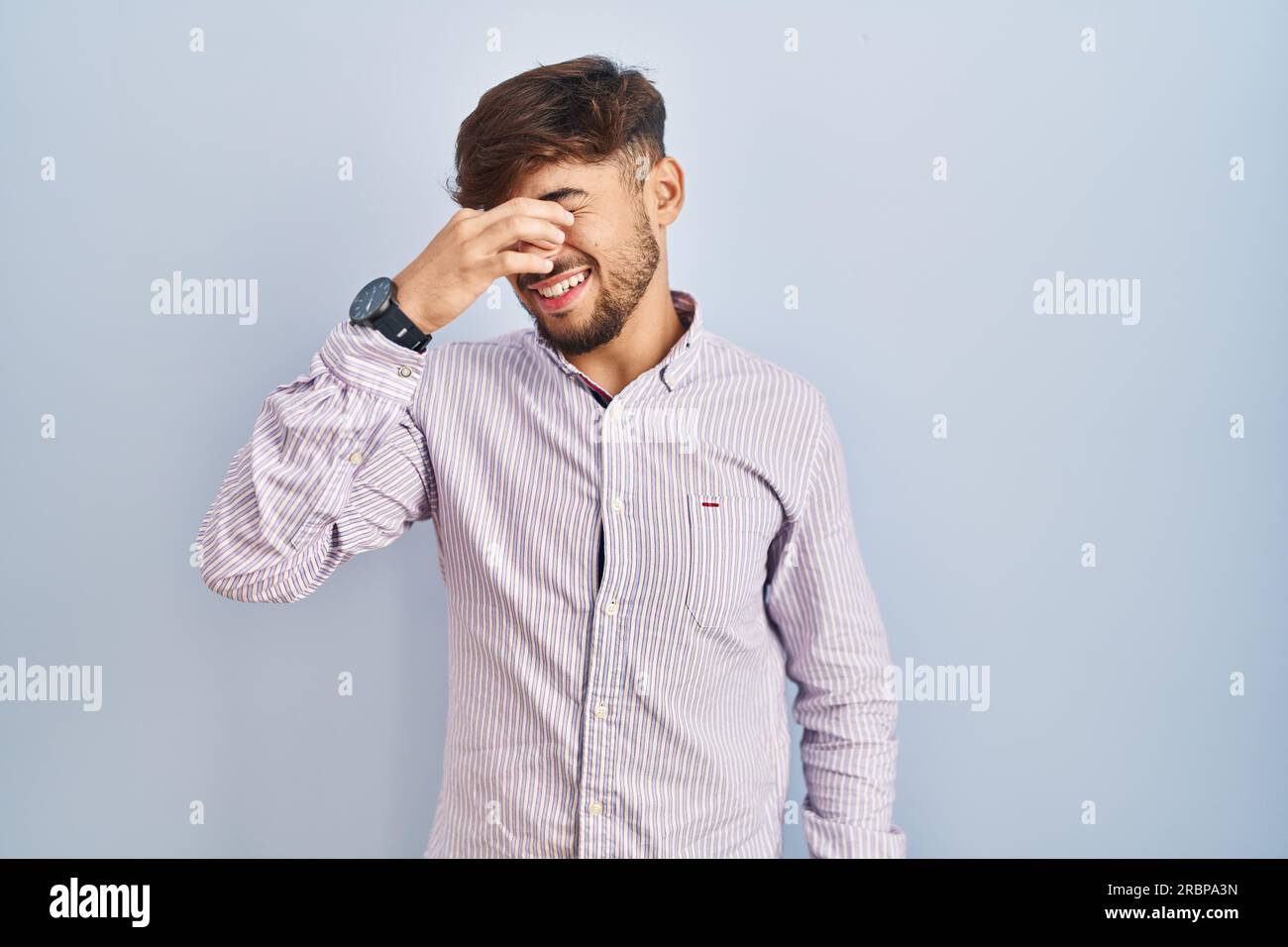 Arab man with beard standing over blue background smelling something ...