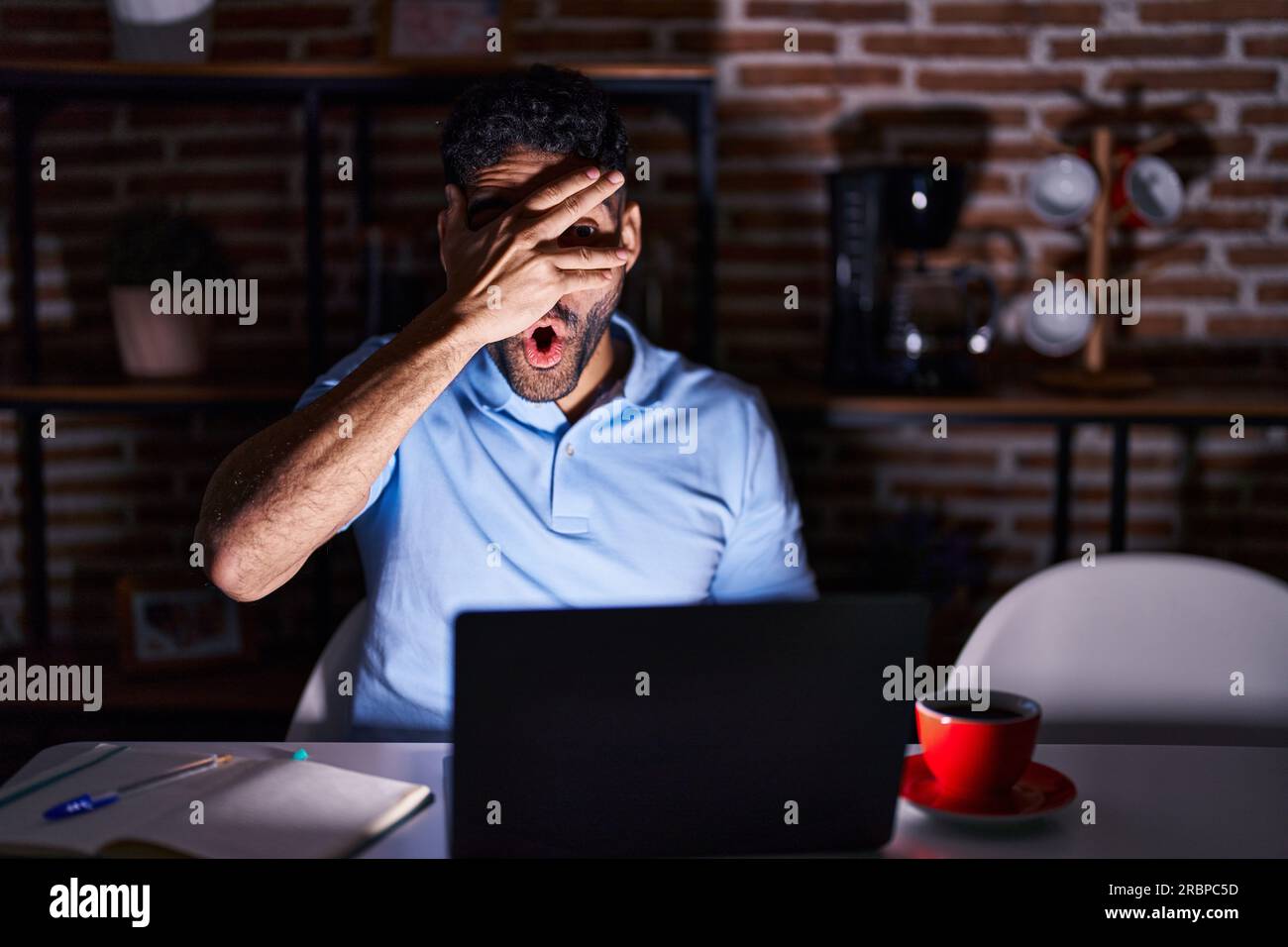 Hispanic man with beard using laptop at night peeking in shock covering ...