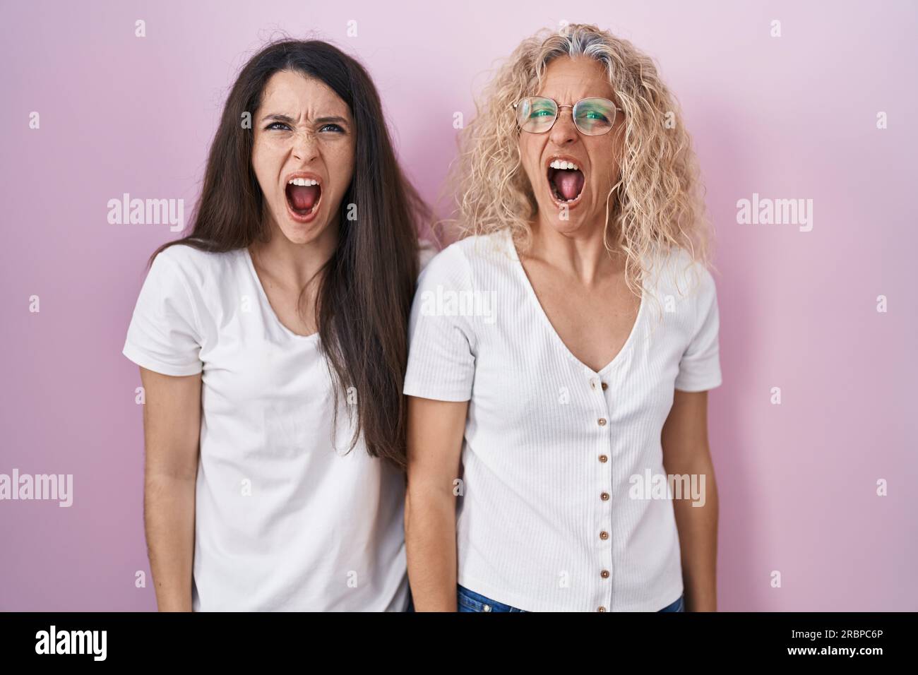 Mother and daughter standing together over pink background angry and ...