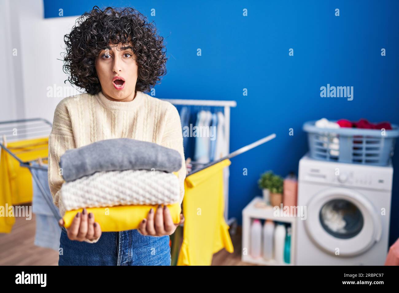 Young brunette woman with curly hair holding clean laundry in shock ...