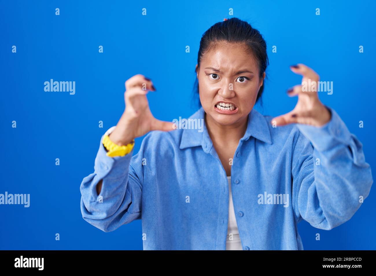 Asian young woman standing over blue background shouting frustrated ...