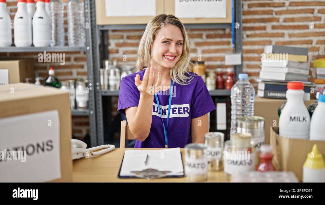 Young blonde woman volunteer sitting on table doing come gesture at ...