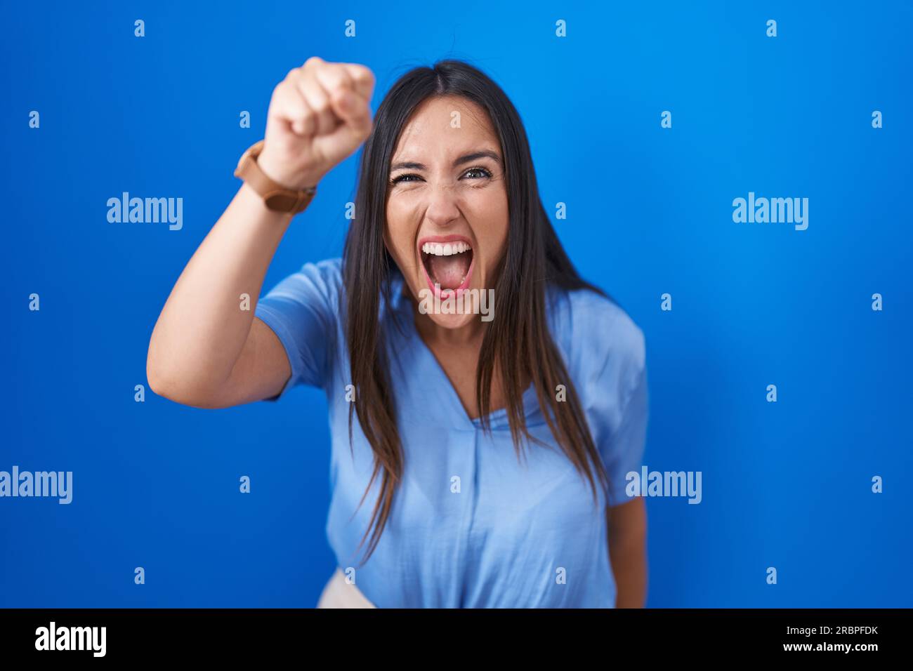Young brunette woman standing over blue background angry and mad ...