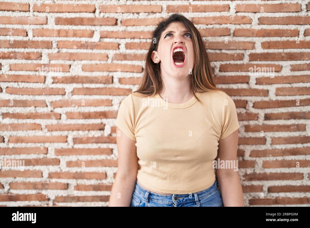 Young brunette woman standing over bricks wall angry and mad screaming frustrated and furious ...