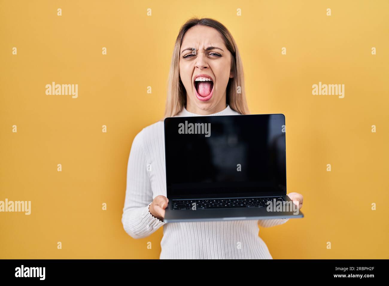 Young blonde woman holding laptop angry and mad screaming frustrated ...