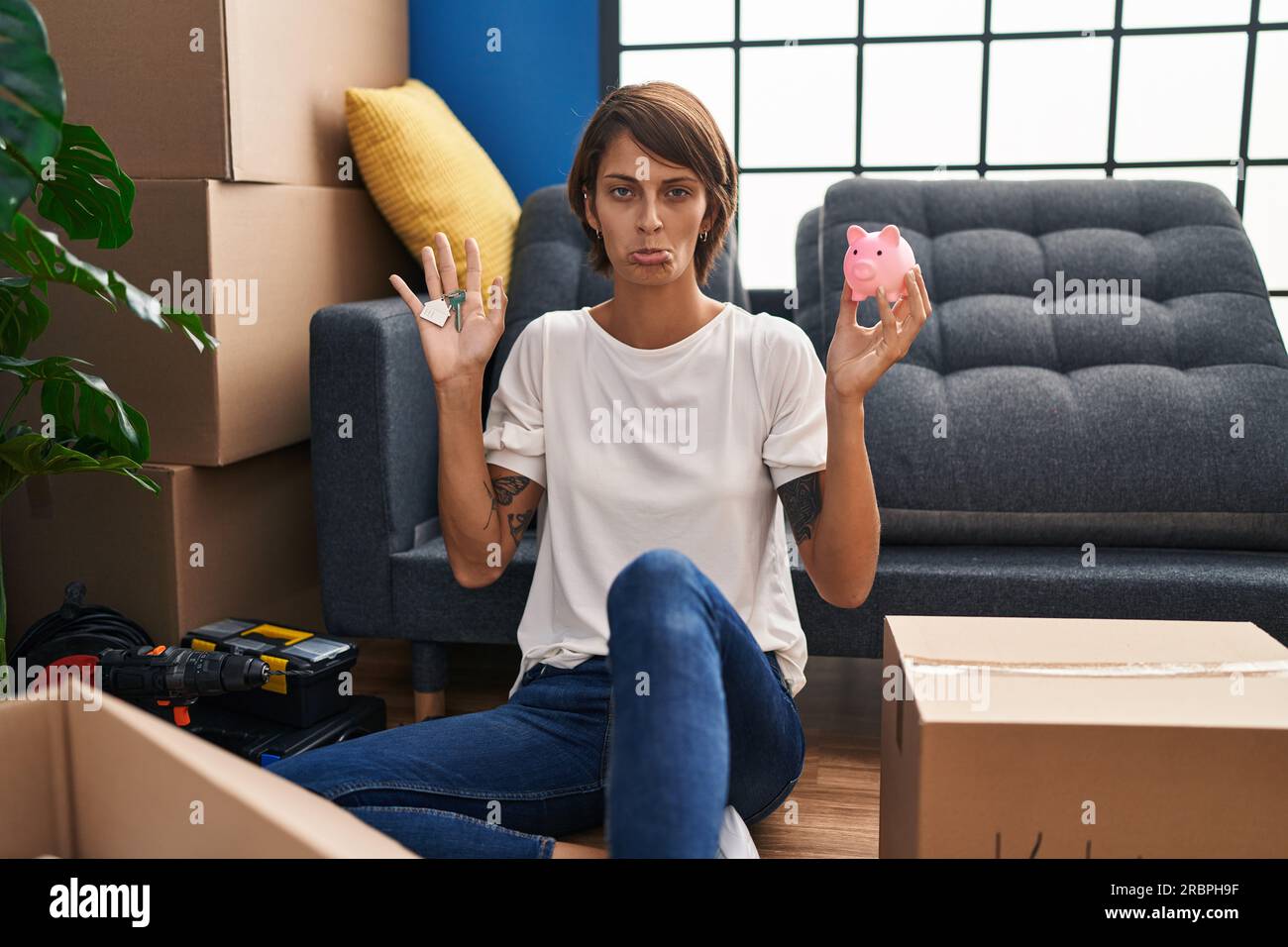 Brunette woman holding keys of new home and piggy bank depressed and ...