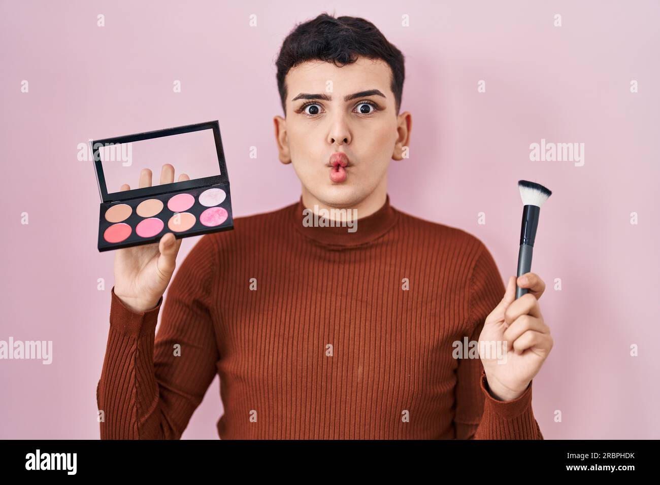 Non binary person holding makeup brush and blush making fish face with ...