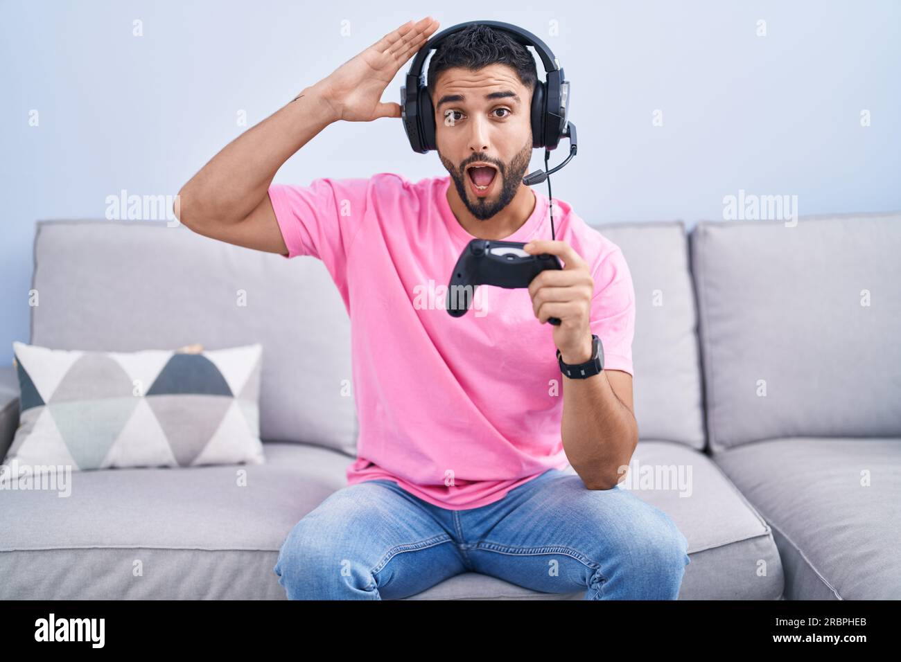 Hispanic young man playing video game holding controller sitting on the sofa crazy and scared ...