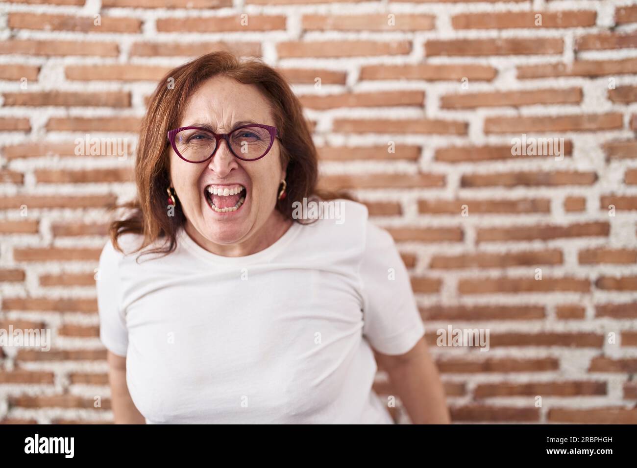 Senior woman with glasses standing over bricks wall angry and mad ...