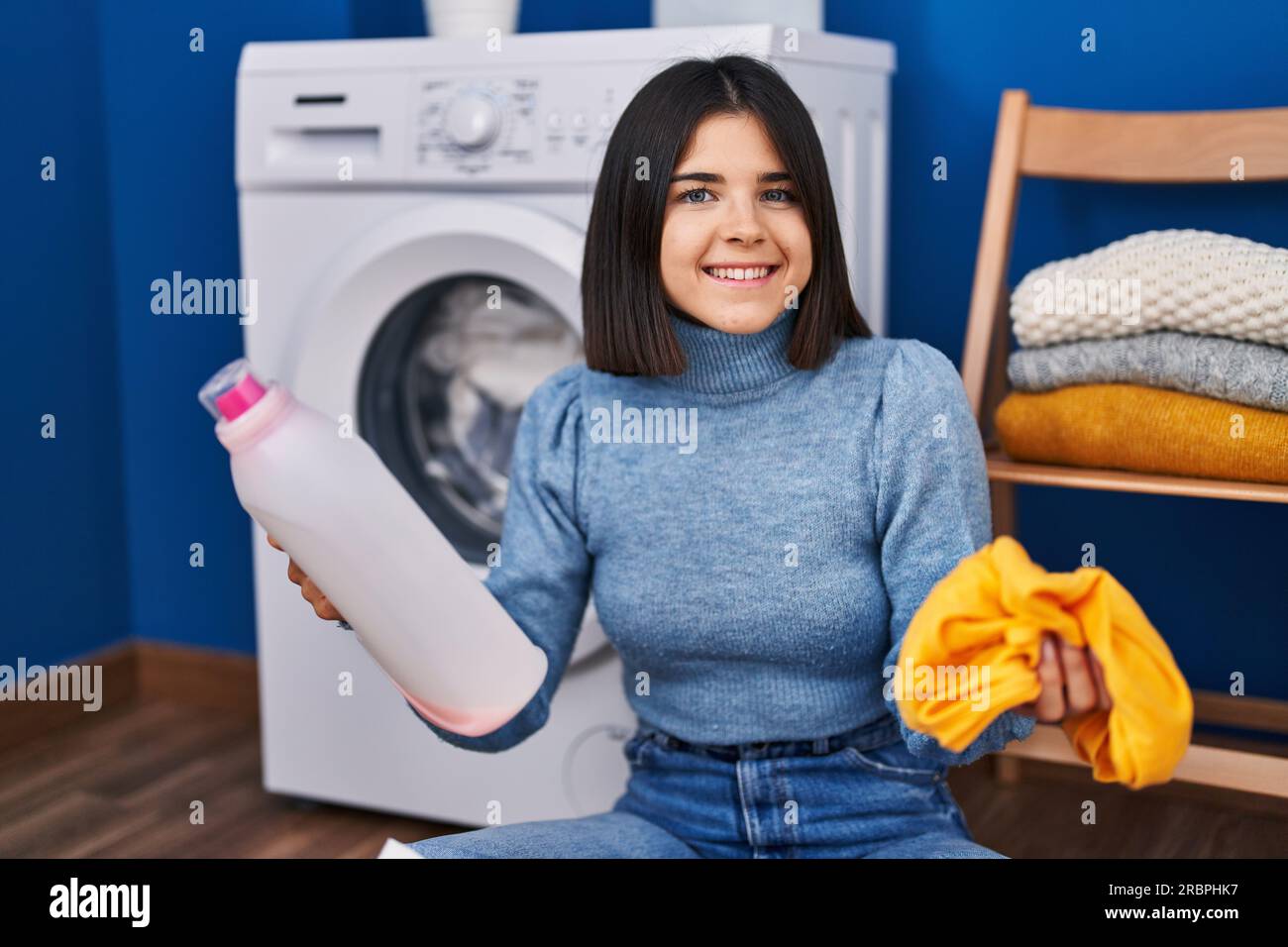 Young beautiful hispanic woman washing clothes holding detergent bottle ...