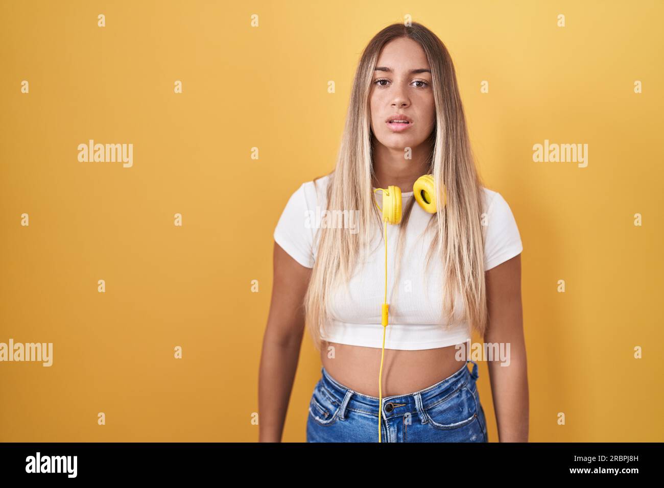 Young blonde woman standing over yellow background wearing headphones ...
