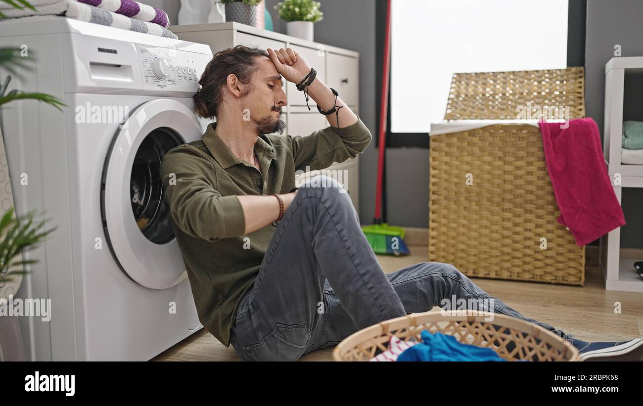 Young hispanic man washing clothes sitting on floor tired at laundry ...