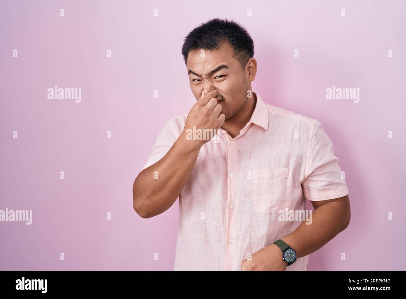 Chinese young man standing over pink background smelling something ...