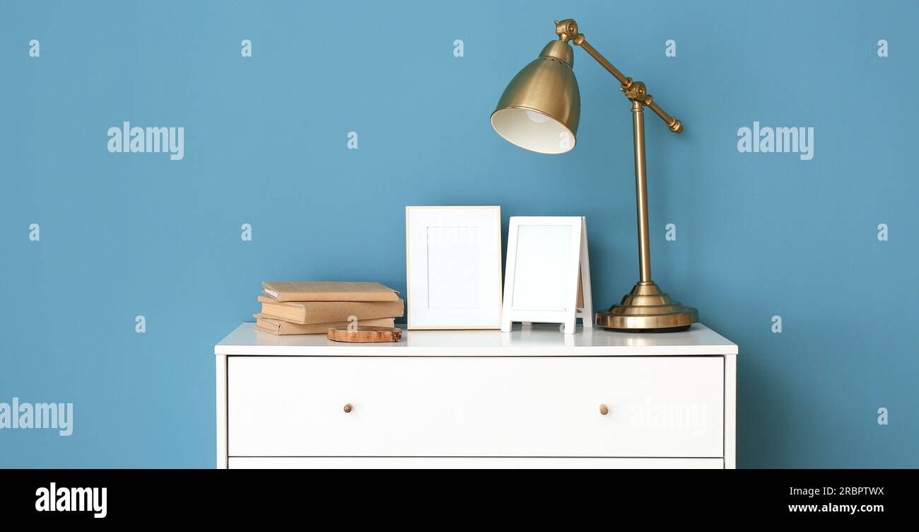 Blank frames, lamp and books on chest of drawers near blue wall. Banner ...