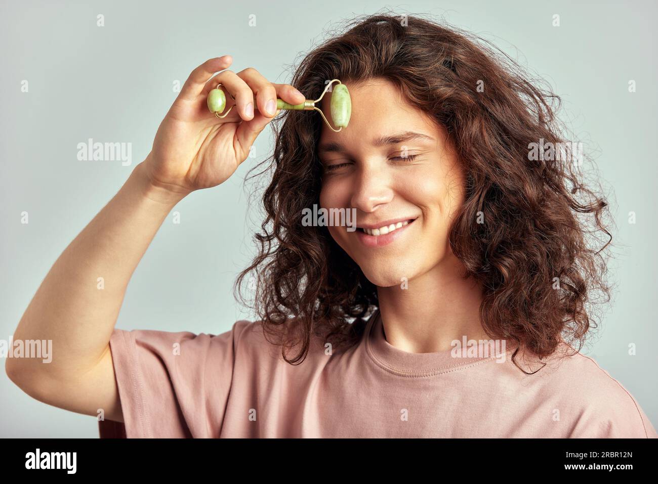 Young handsome long haired guy using jade de-puffing face roller to ...