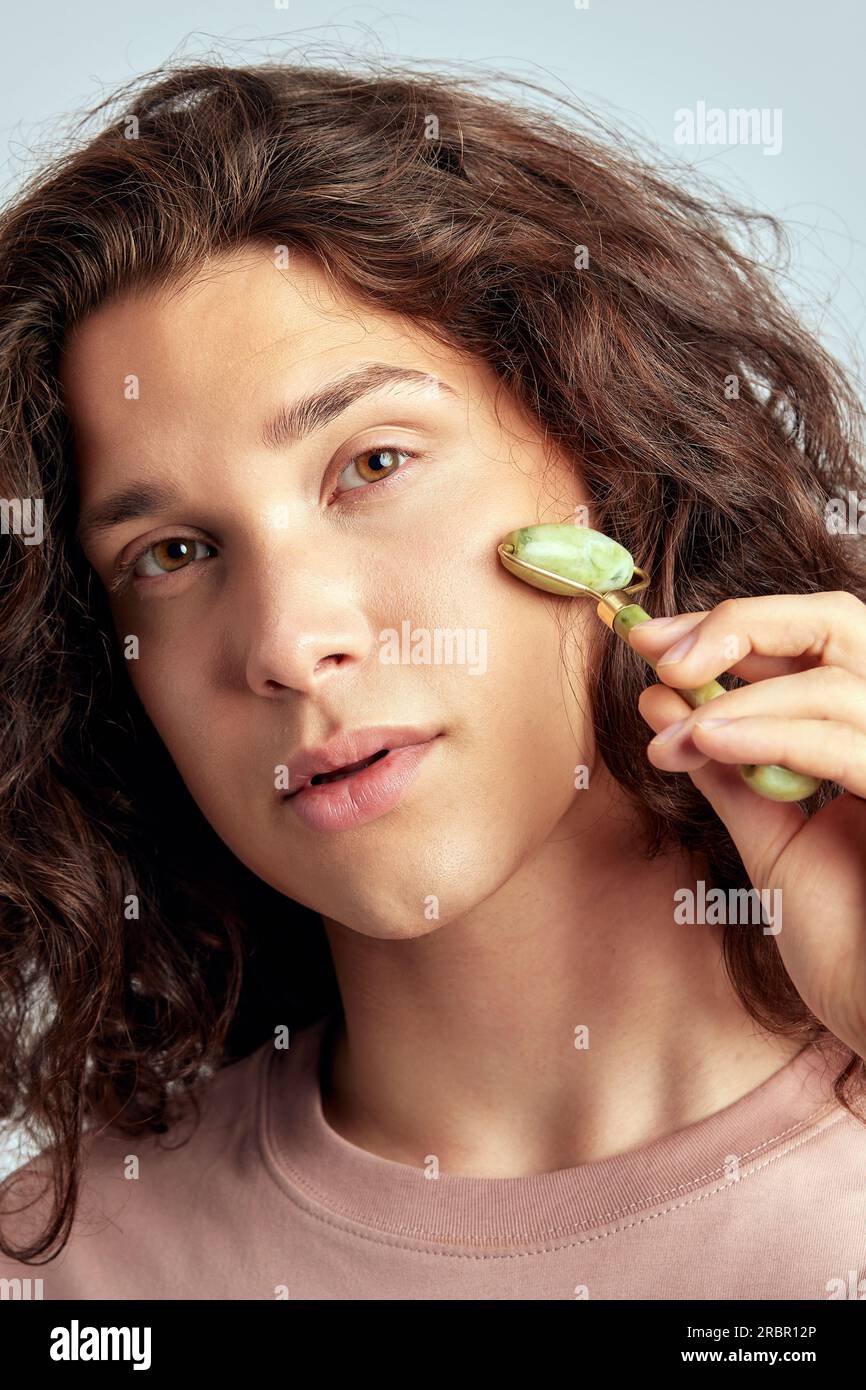 Young handsome long haired guy using jade de-puffing face roller to ...