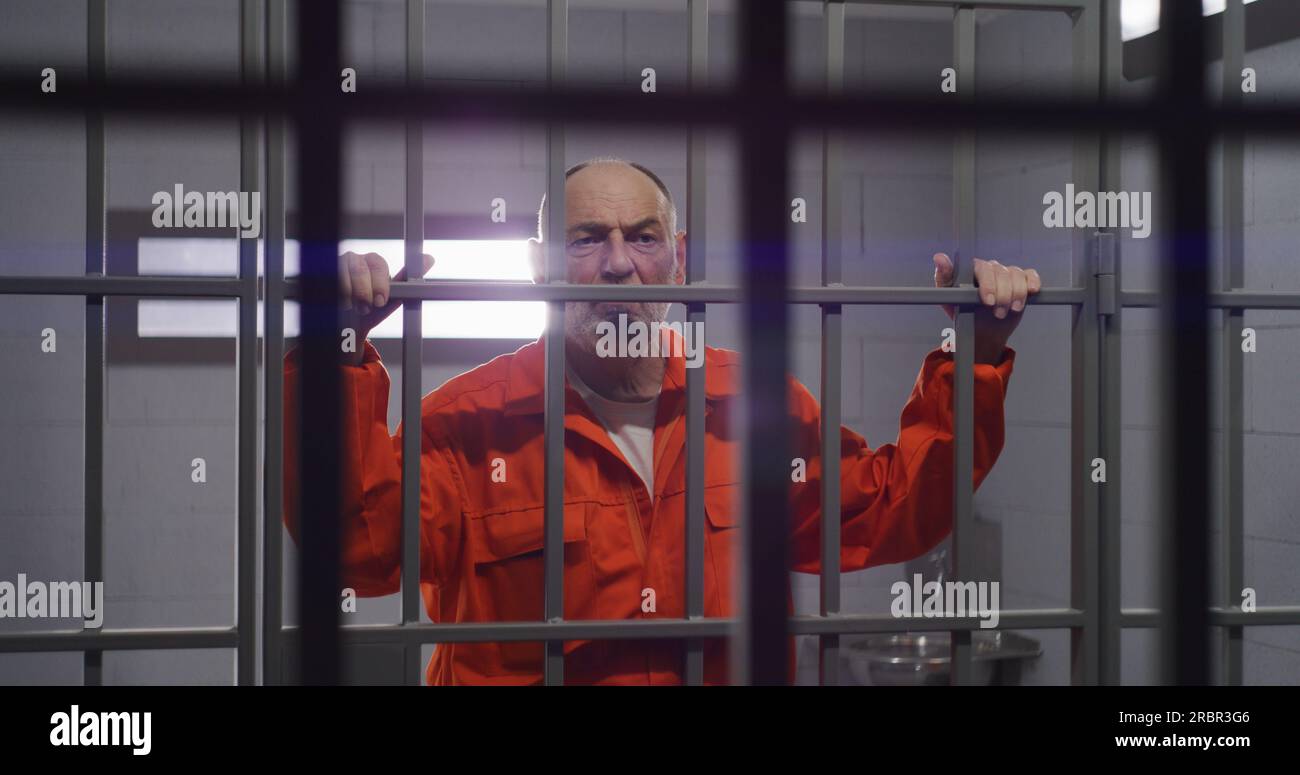 Elderly prisoner in orange uniform holds hands on metal bars. Criminal ...