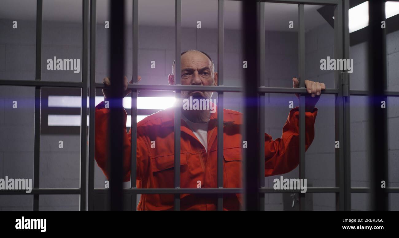 Elderly prisoner in orange uniform holds hands on metal bars. Criminal ...