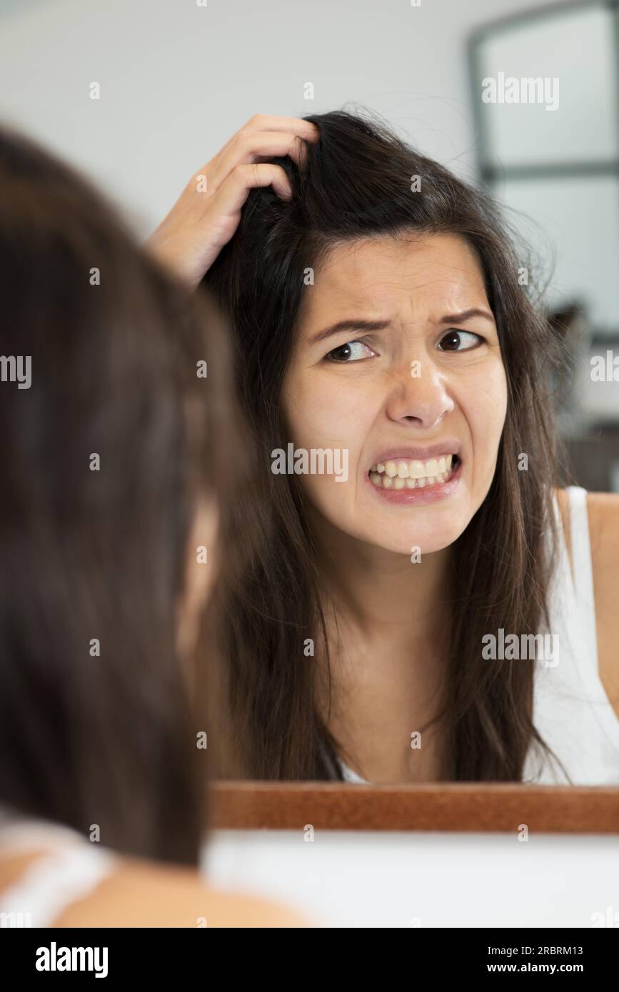 Woman having a bad hair day grimacing in disgust as she looks in the mirror and runs her hands ...