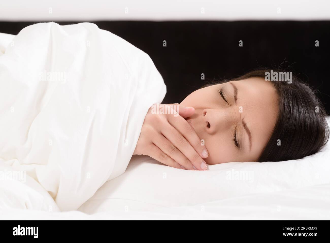 Tired attractive young woman lying in bed yawning with her hand to her mouth and eyes closed as ...