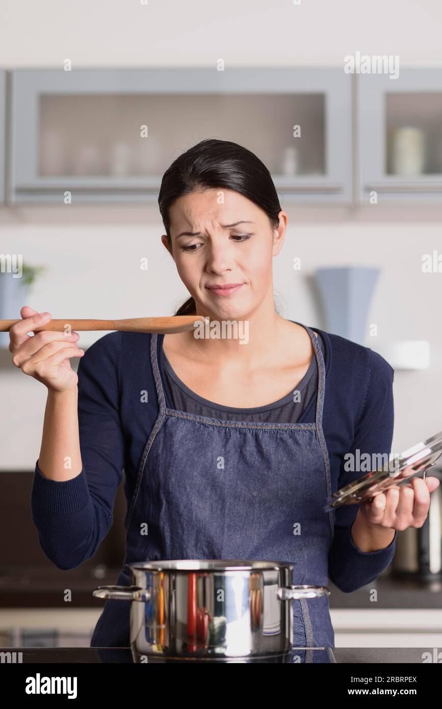 Attractive young female cook standing at the hob in her apron tasting ...