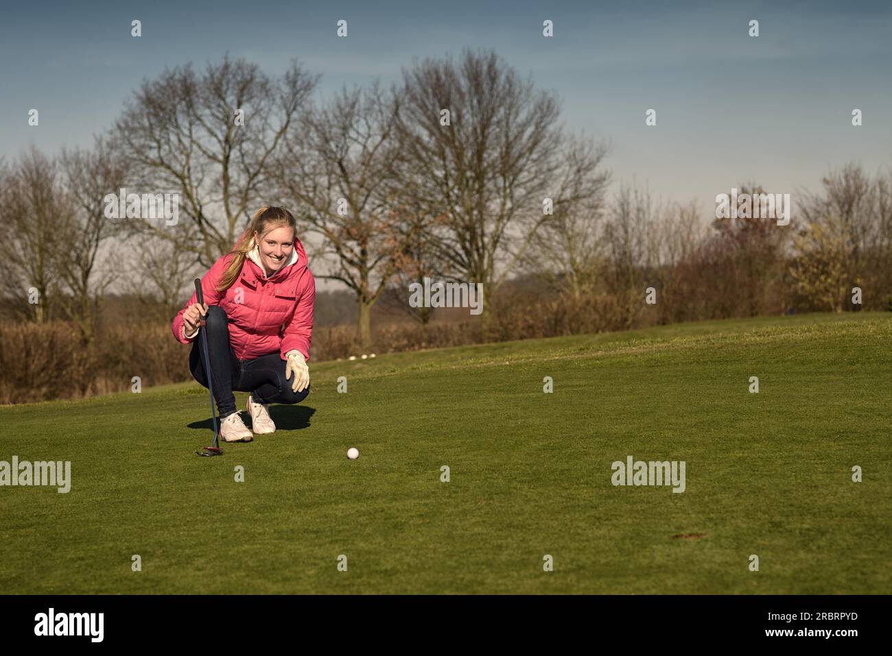 Female golfer lining up a putt kneeling on the green looking at the hole to check the grass and ...