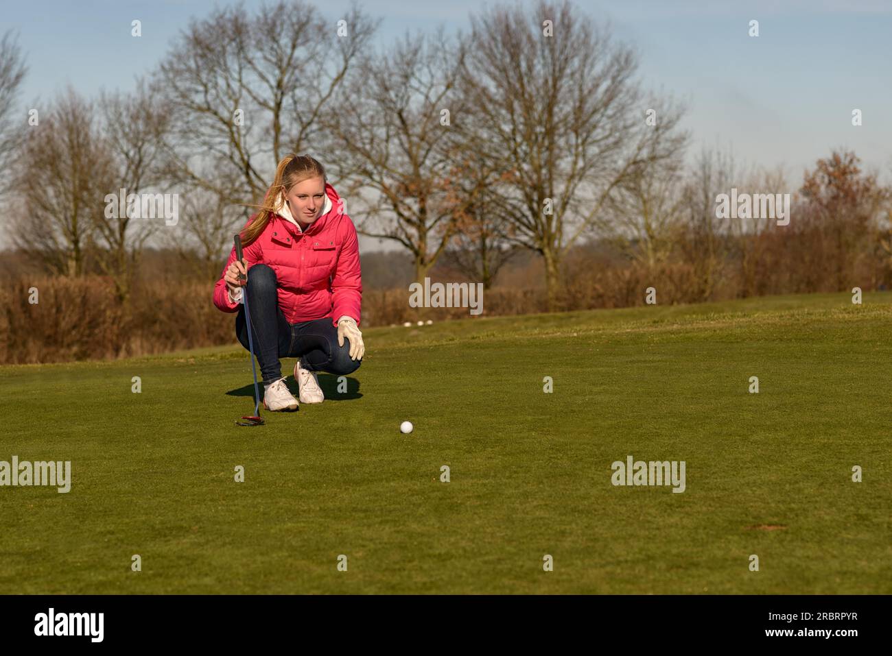 Female golfer lining up a putt kneeling on the green looking at the hole to check the grass and ...