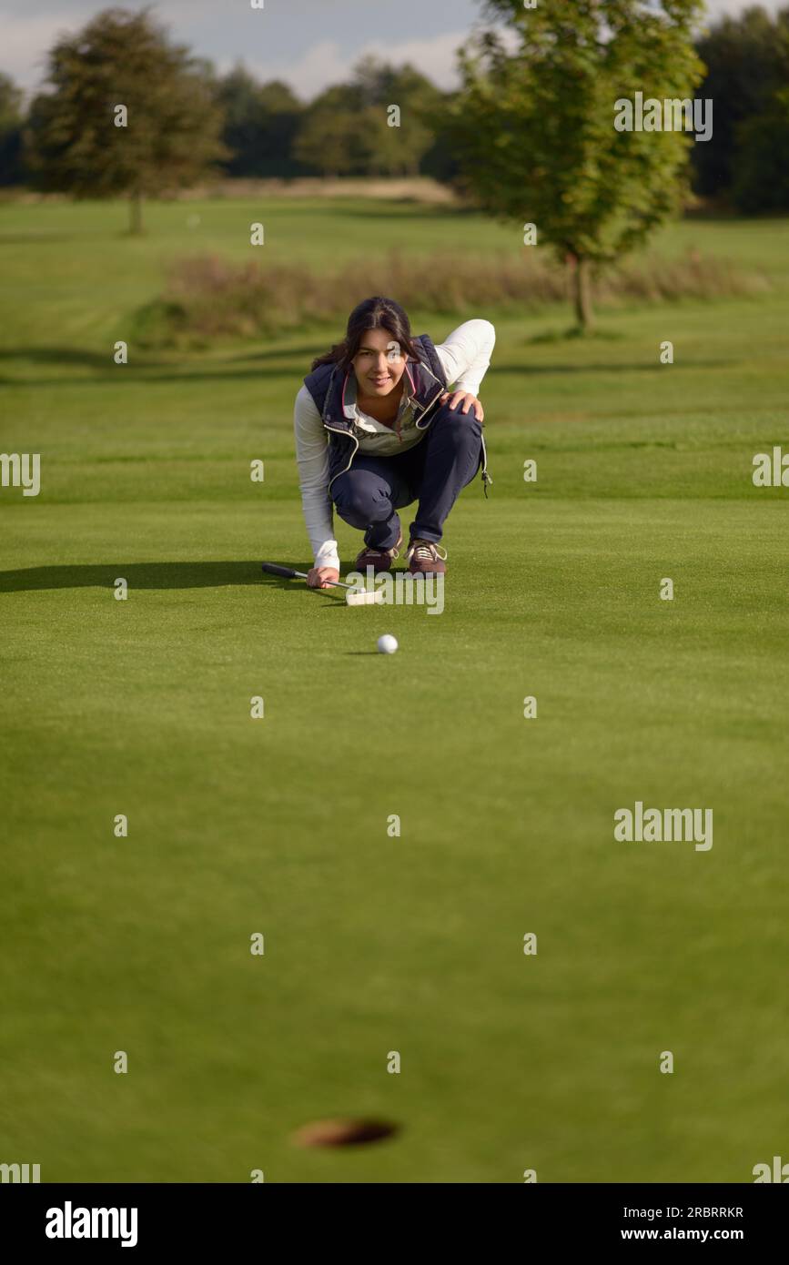 Female golfer lining up a putt kneeling on the green looking at the hole to check the grass and ...