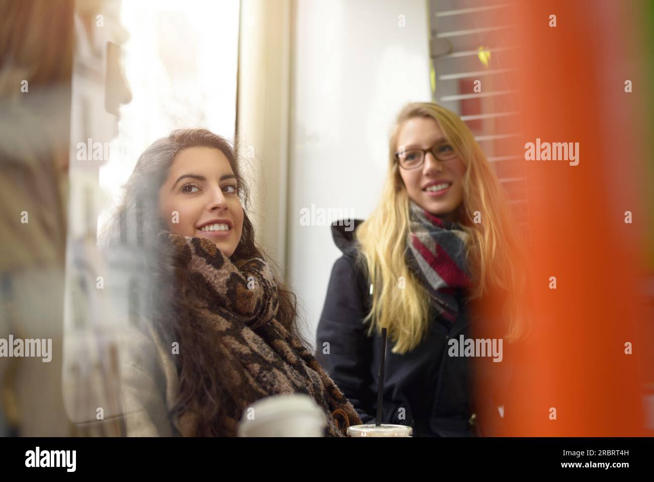 Blond and brunette female friends wearing coats talk in public space ...