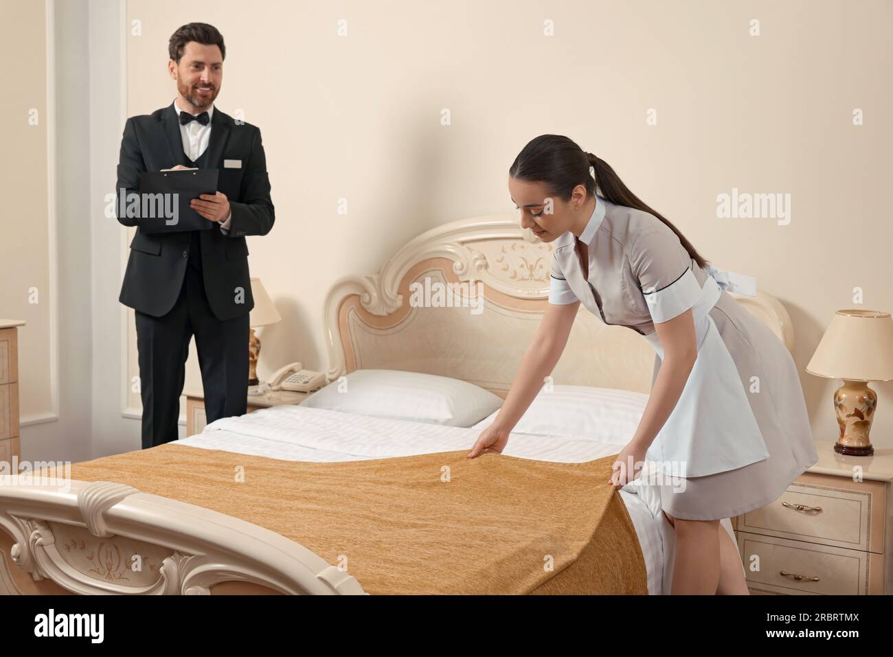 Man wearing suit with clipboard checking maid's work in hotel room ...
