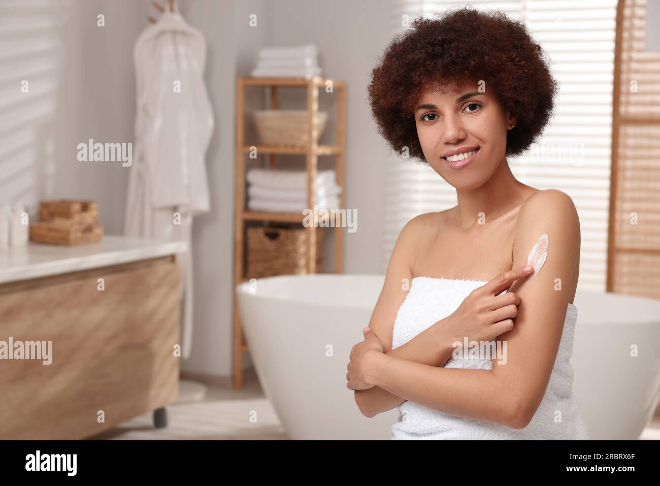 Beautiful young woman applying body cream onto arm in bathroom, space ...