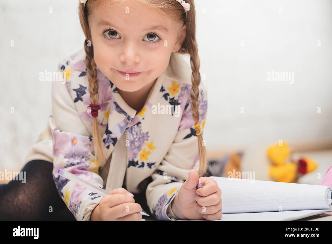 A little girl with her hair up and a headband, sits drawing. She pauses ...