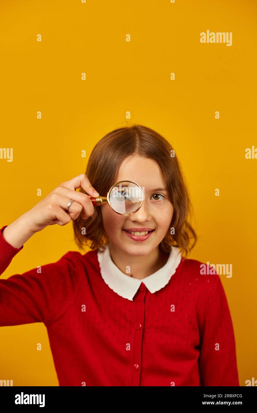 Stylish smiling school teenage girl holding magnifying glass on her eye ...