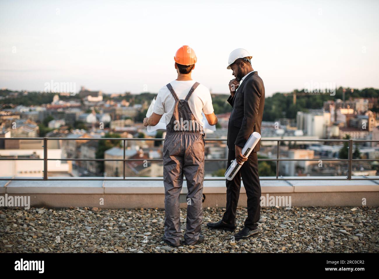 Back view of two male developers wearing hardhats standing on rooftop ...