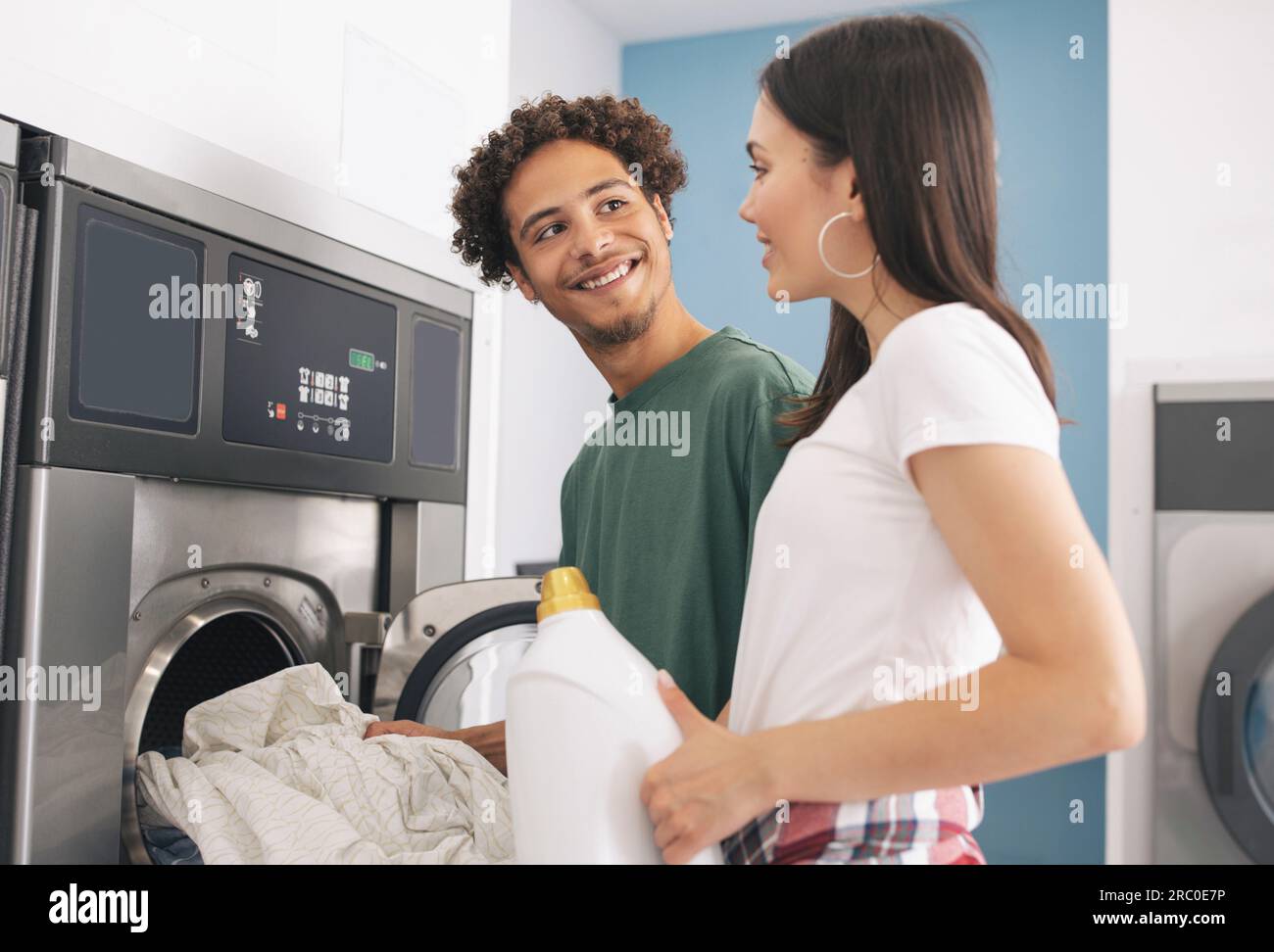Happy couple putting laundry into washing machine at public laundromat ...