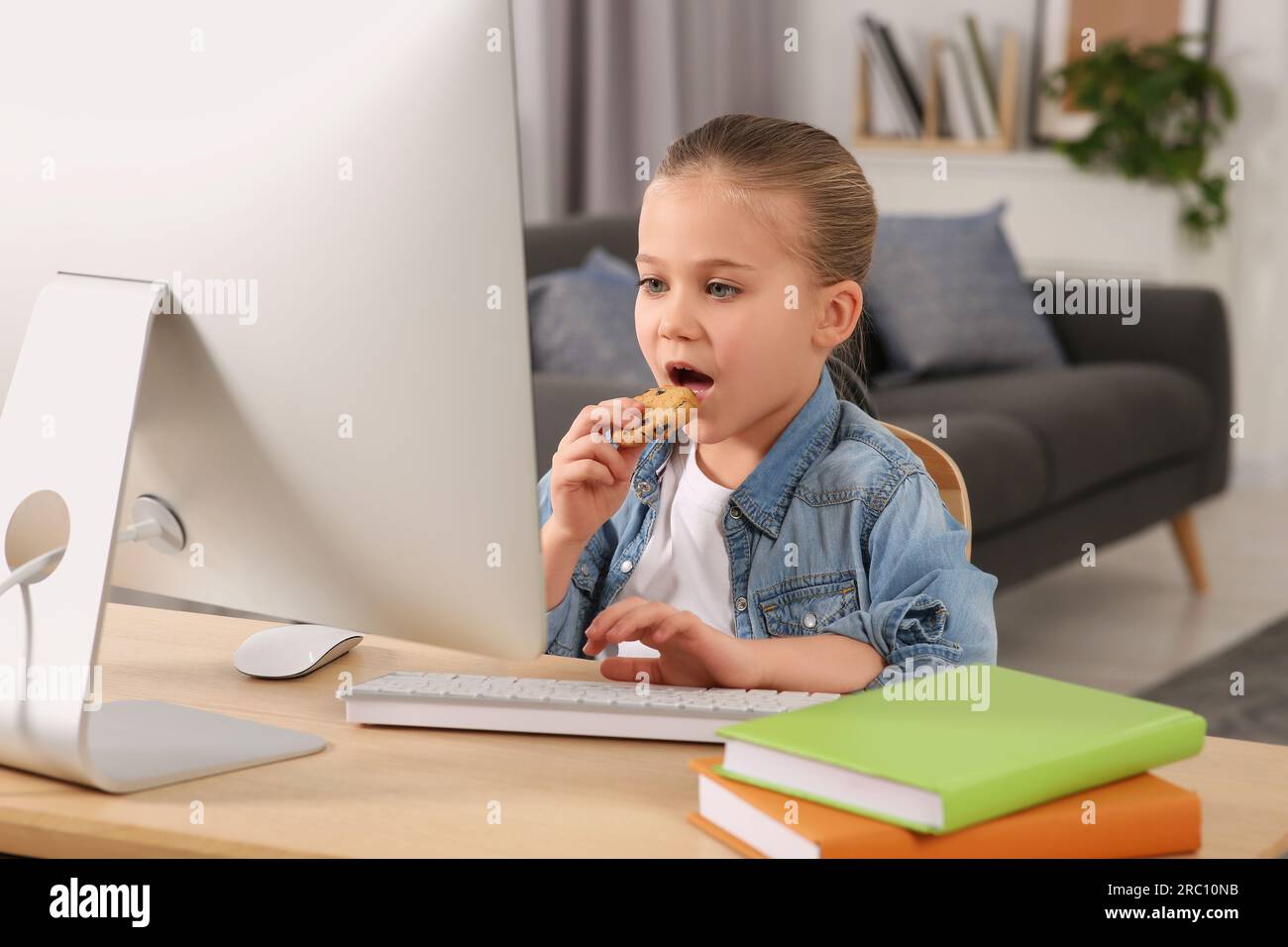 Little girl eating biscuit while using computer at table in room ...