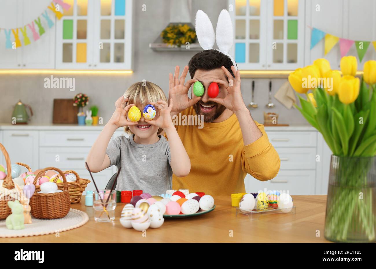 Father and his cute son covering eyes with Easter eggs at table in ...