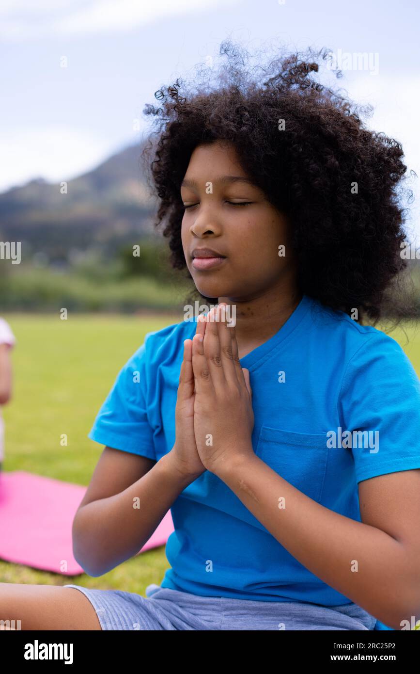Happy african american schoolboy doing yoga and meditating on field at ...