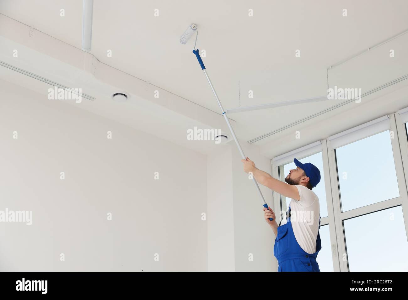 Worker in uniform painting ceiling with roller indoors. Space for text ...
