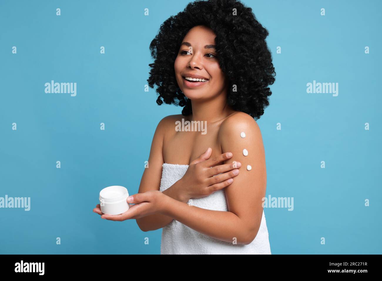 Young woman applying body cream onto shoulder on light blue background ...