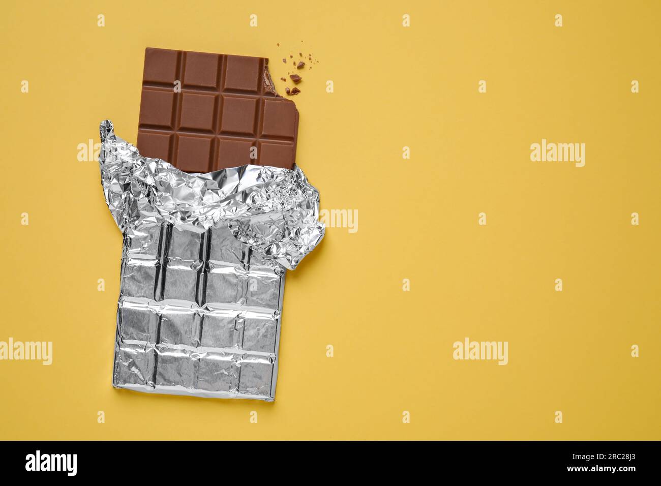 Bitten milk chocolate bar wrapped in foil on pale yellow background ...