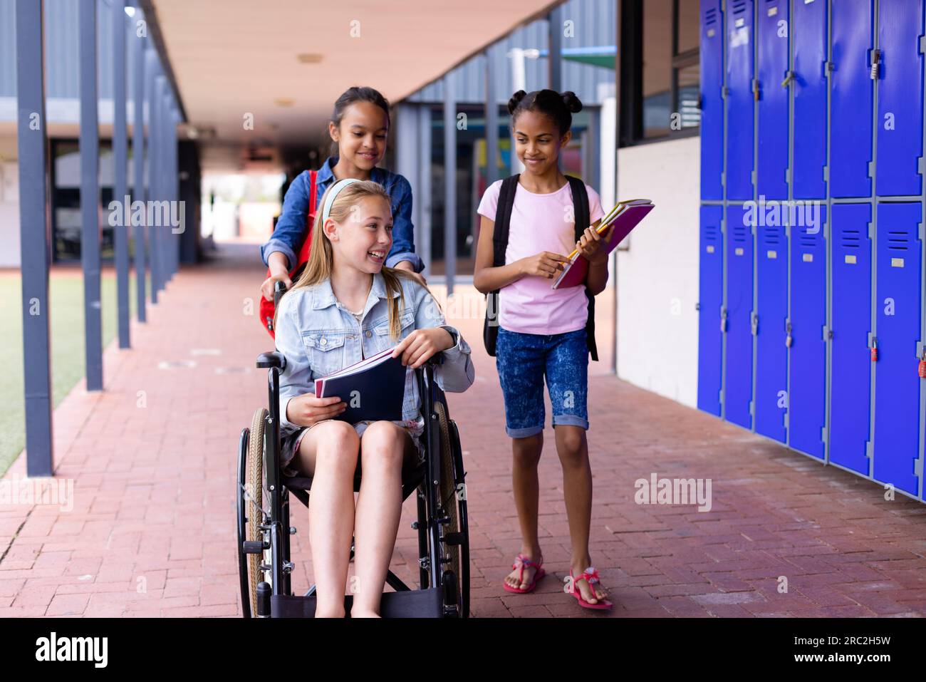 Happy diverse schoolgirl in wheelchair with her friends in corridor at ...