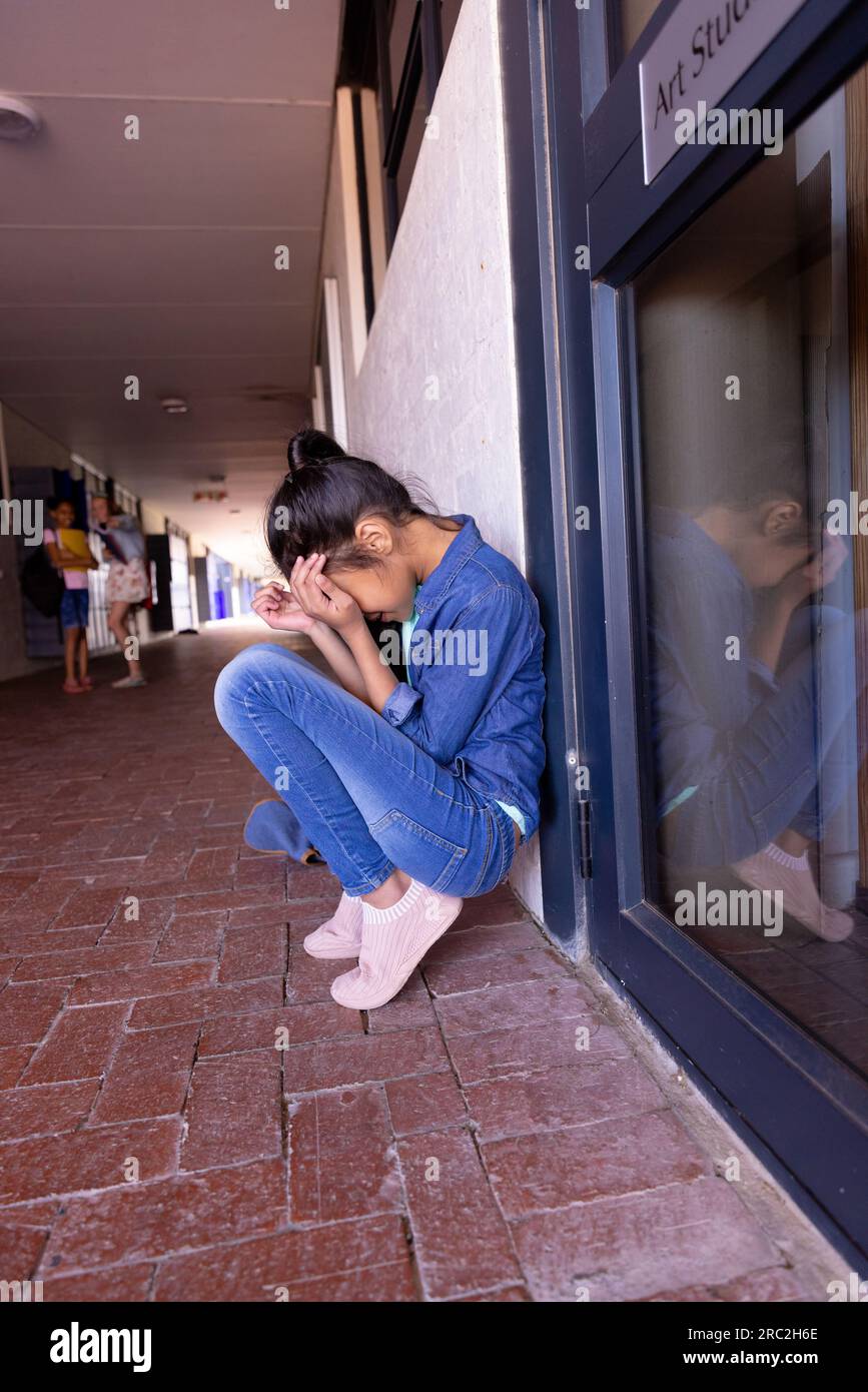 Sad biracial schoolgirl sitting in corridor with diverse schoolchildren in background Stock ...