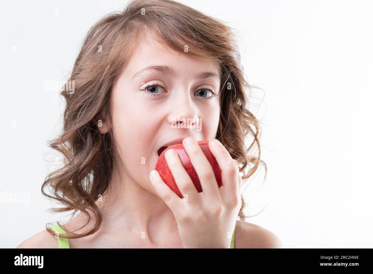 A curly-haired girl in a green tank top bites into a juicy red apple ...