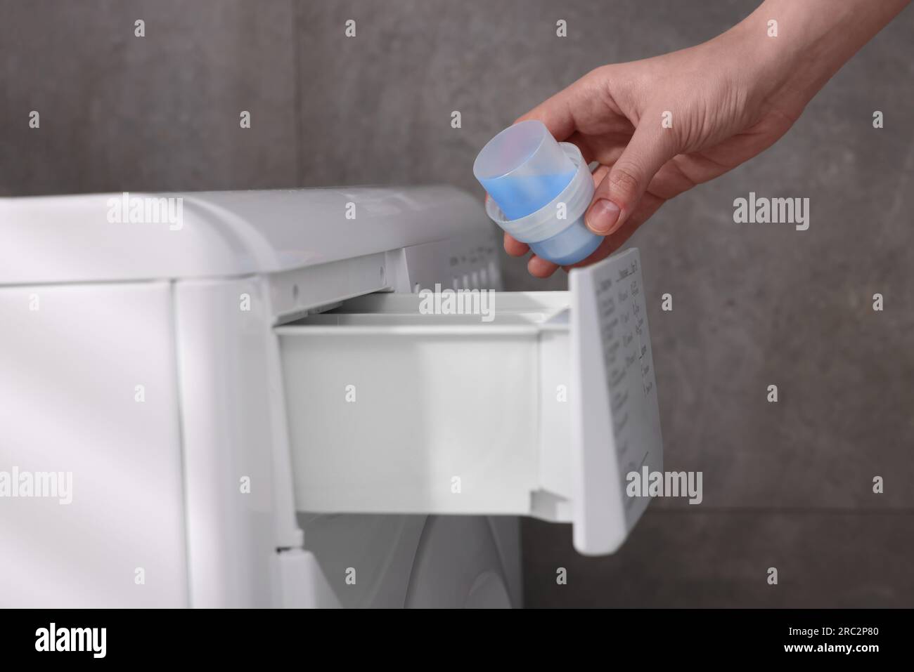 Woman pouring fabric softener from cap into washing machine near grey ...