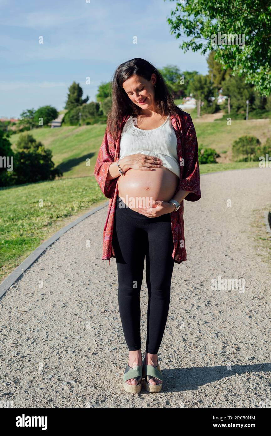 Full body of glad expecting female in maternity clothes touching gently ...