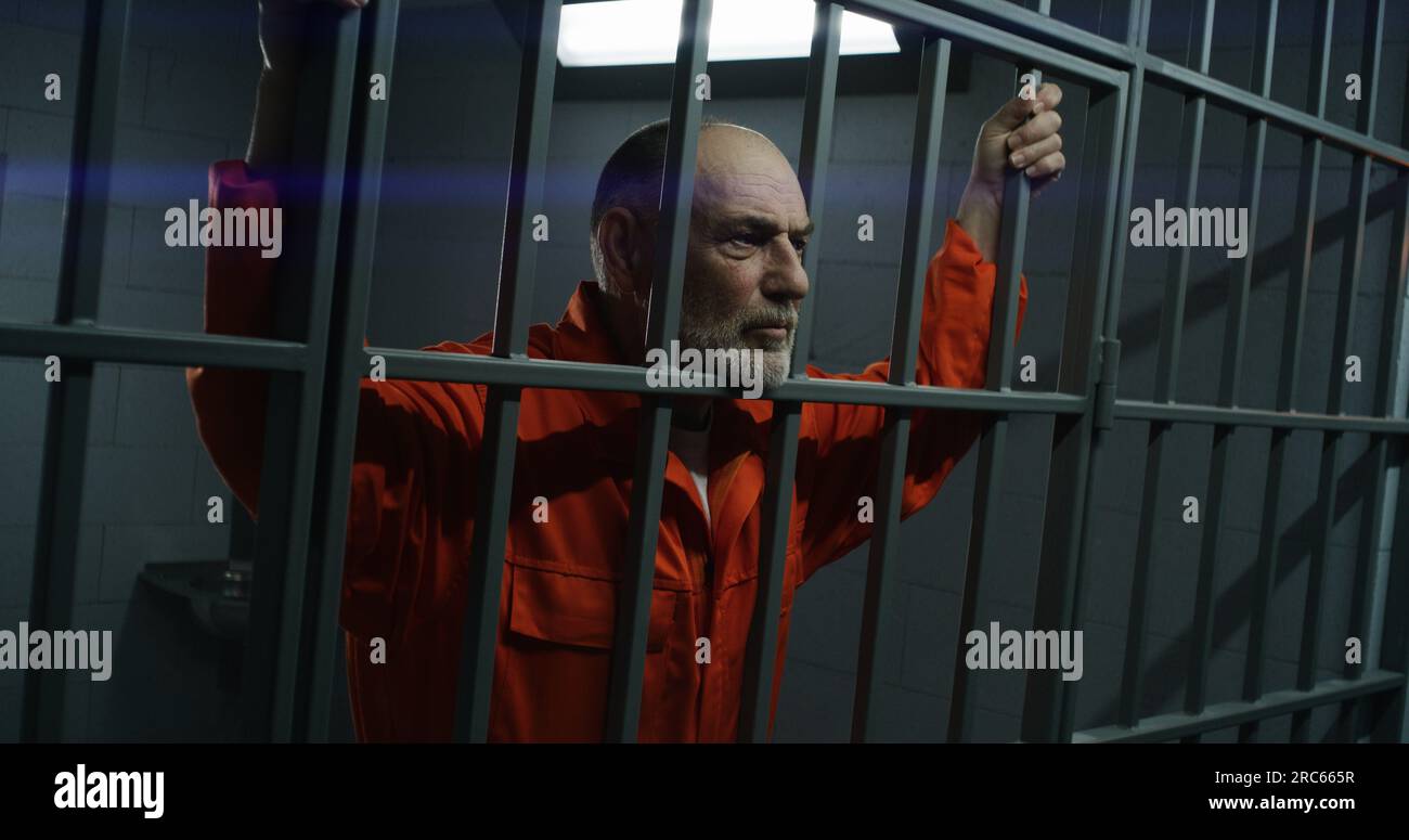 Elderly prisoner in orange uniform stretches fingers, leans on metal ...