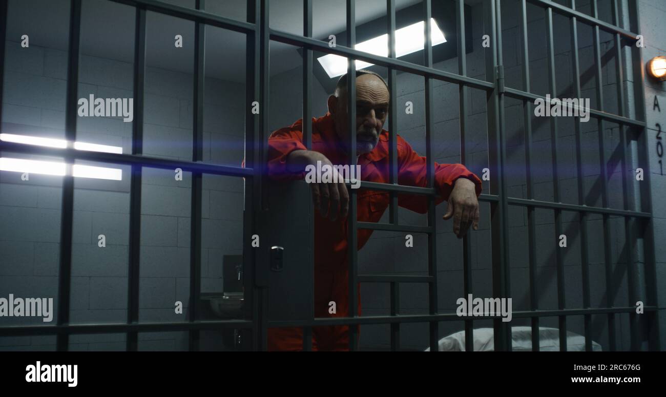 Elderly prisoner in orange uniform leans hands on metal bars. Criminal ...